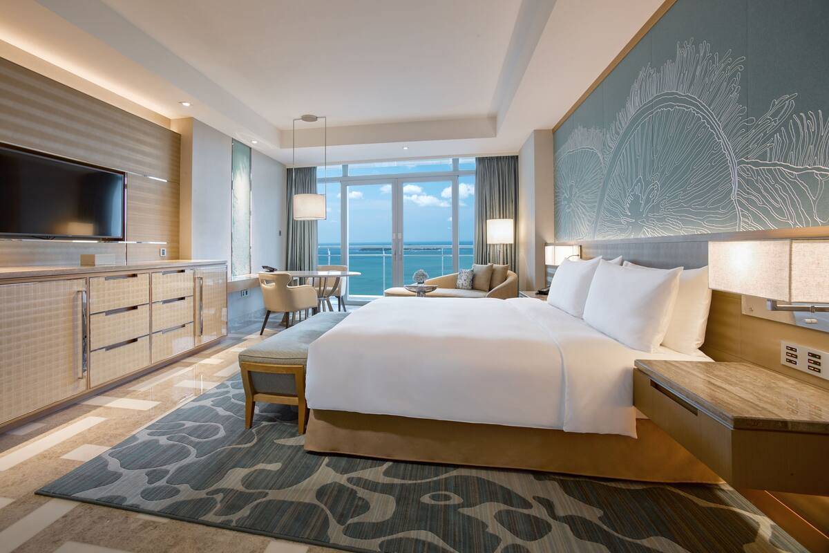 Quarto de hotel moderno com uma cama grande, televisão de tela plana, escrivaninha e vista para o oceano.