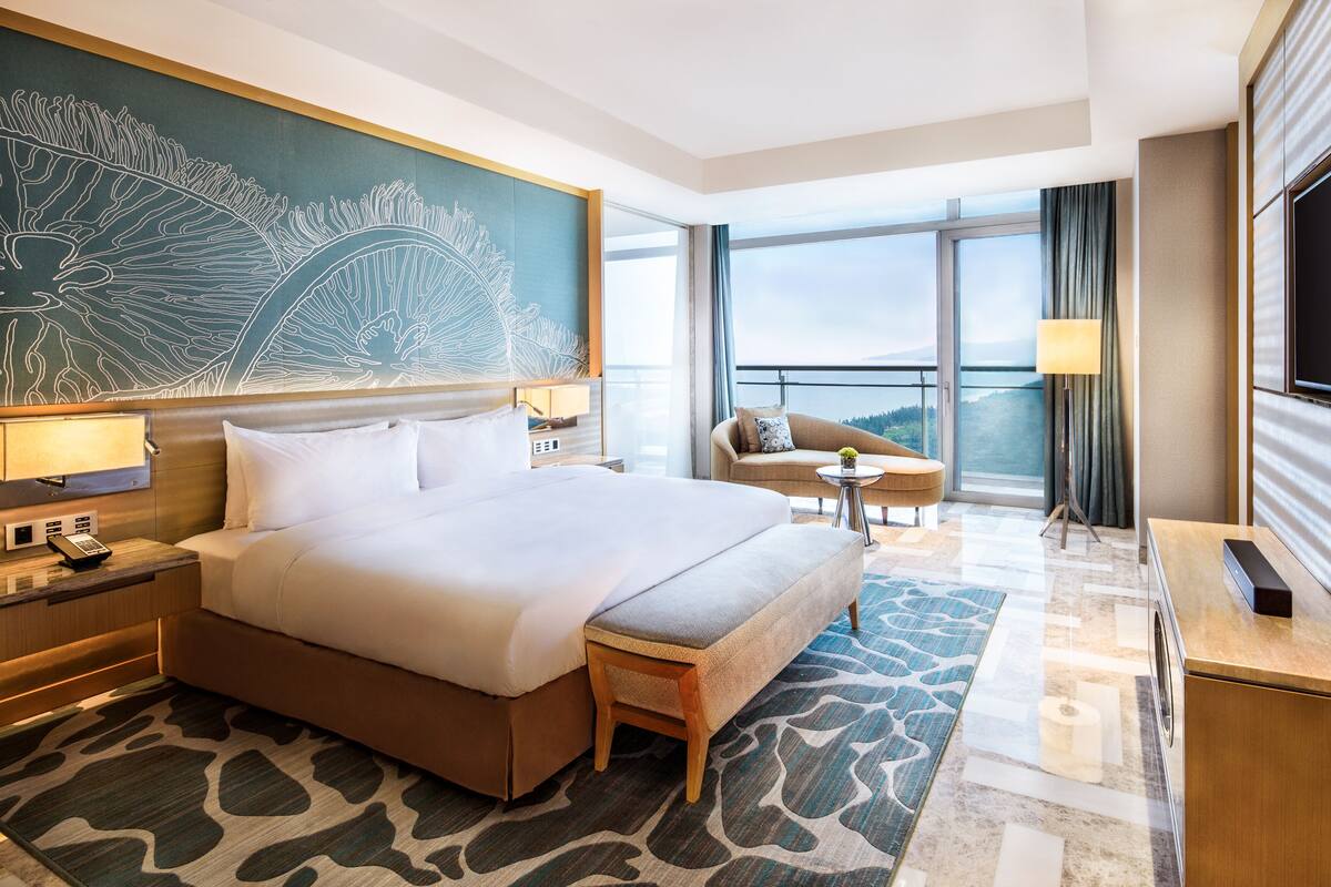 Quarto de hotel com uma cama grande, um sofá, uma mesinha, uma televisão e vista para o exterior.