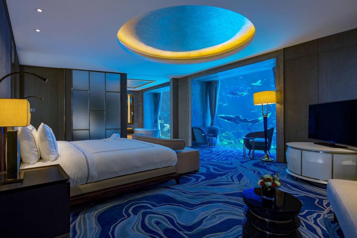 Quarto de hotel com cama, escrivaninha, televisão, minibar e vista para um aquário.