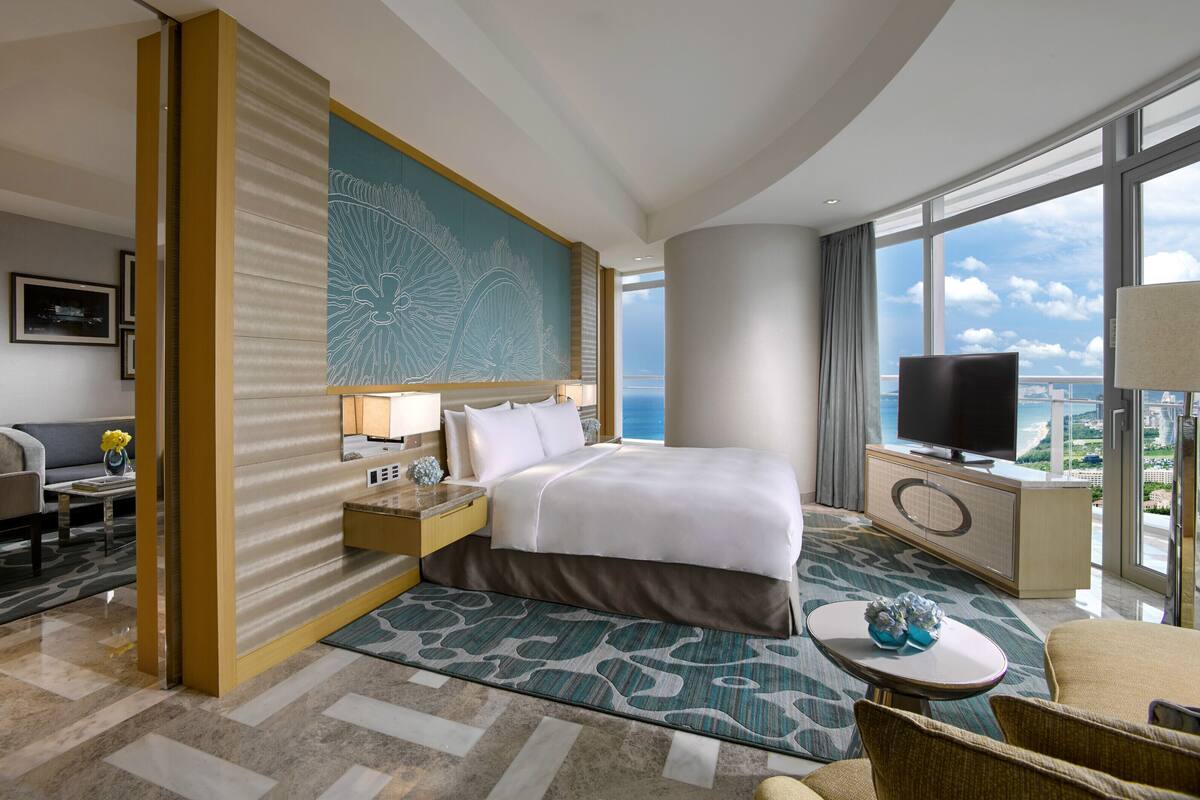 Quarto de hotel com uma cama grande, televisão, escrivaninha com luminária, cadeira e vista para o oceano, além de uma janela ampla.