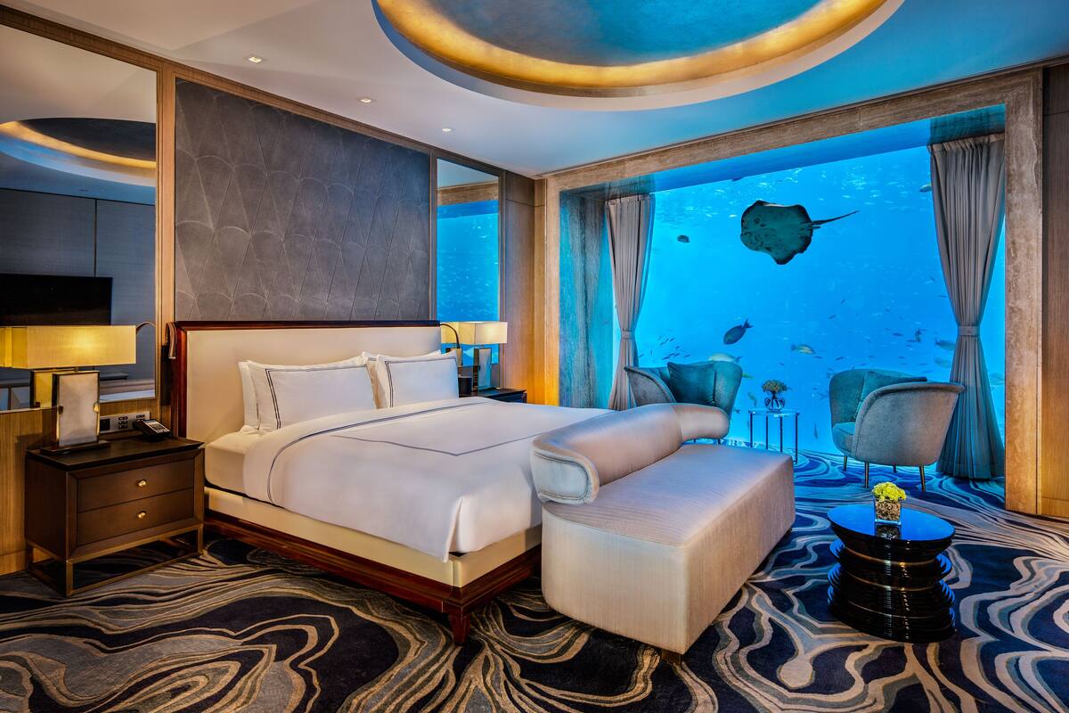 Quarto de hotel com cama, área de estar, televisão e grande janela com vista subaquática.
