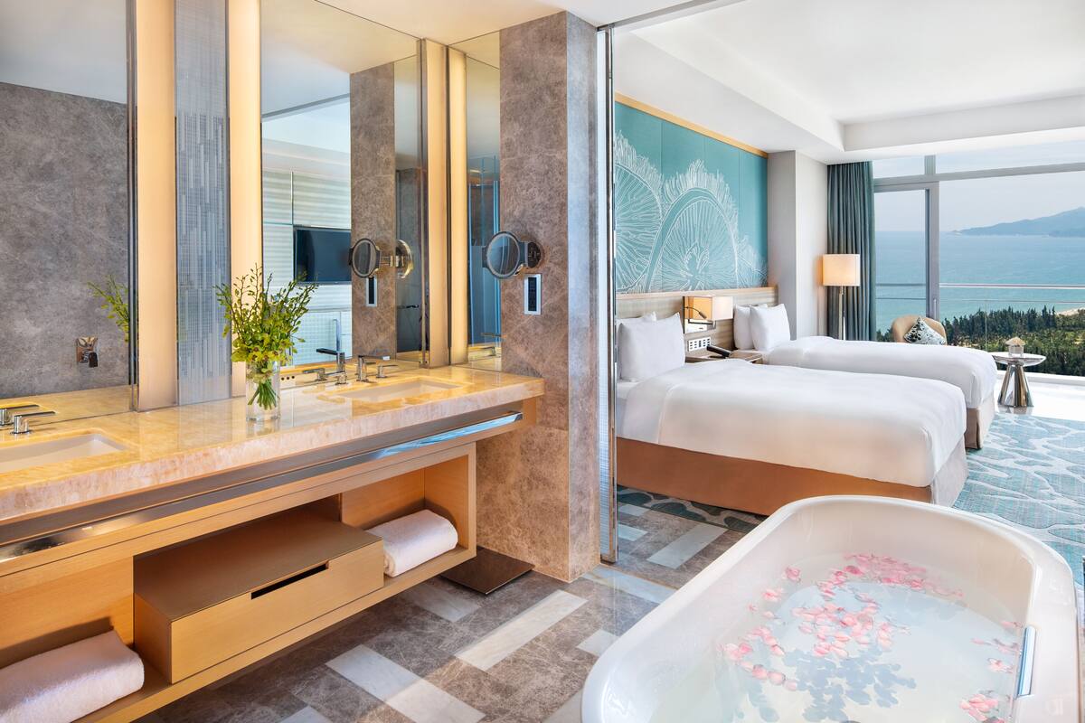 Quarto de hotel moderno com banheiro amplo, cama com duas almofadas, banheira com design floral e vista para o mar.