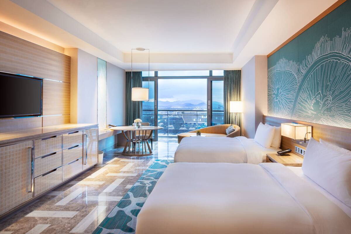 Quarto de hotel com duas camas, uma escrivaninha e uma janela grande com vista para a cidade.