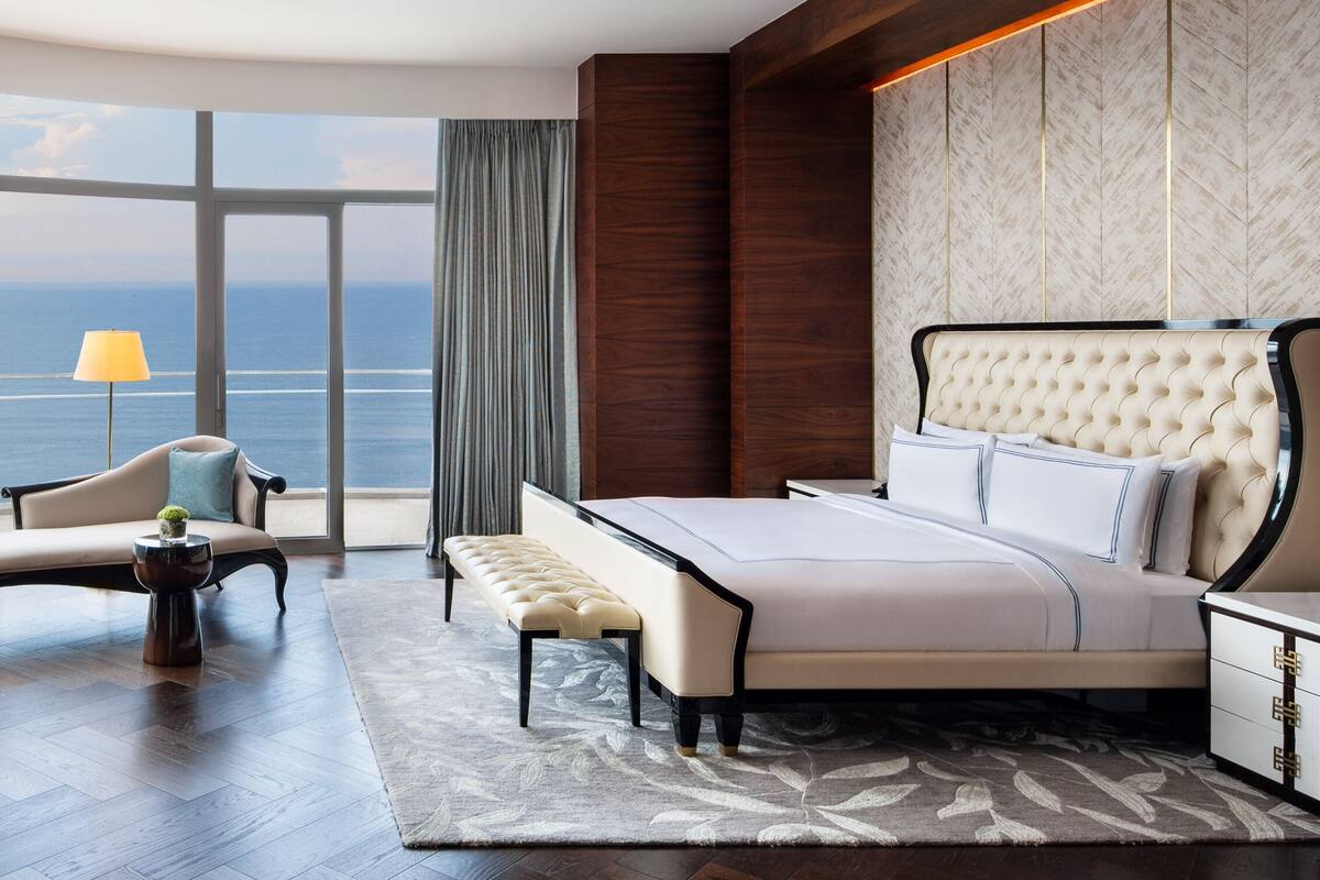 Quarto de hotel moderno com uma janela ampla que oferece vista para o mar, televisão de tela plana, sofá, cama e uma pequena mesa lateral.