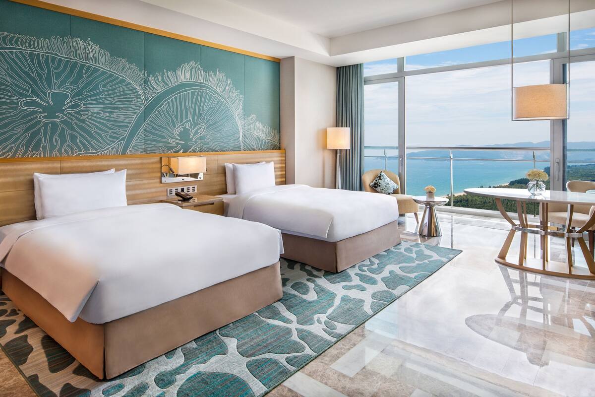 Quarto de hotel com duas camas, uma janela ampla e uma vista, área de refeições e um objeto decorativo na parede.