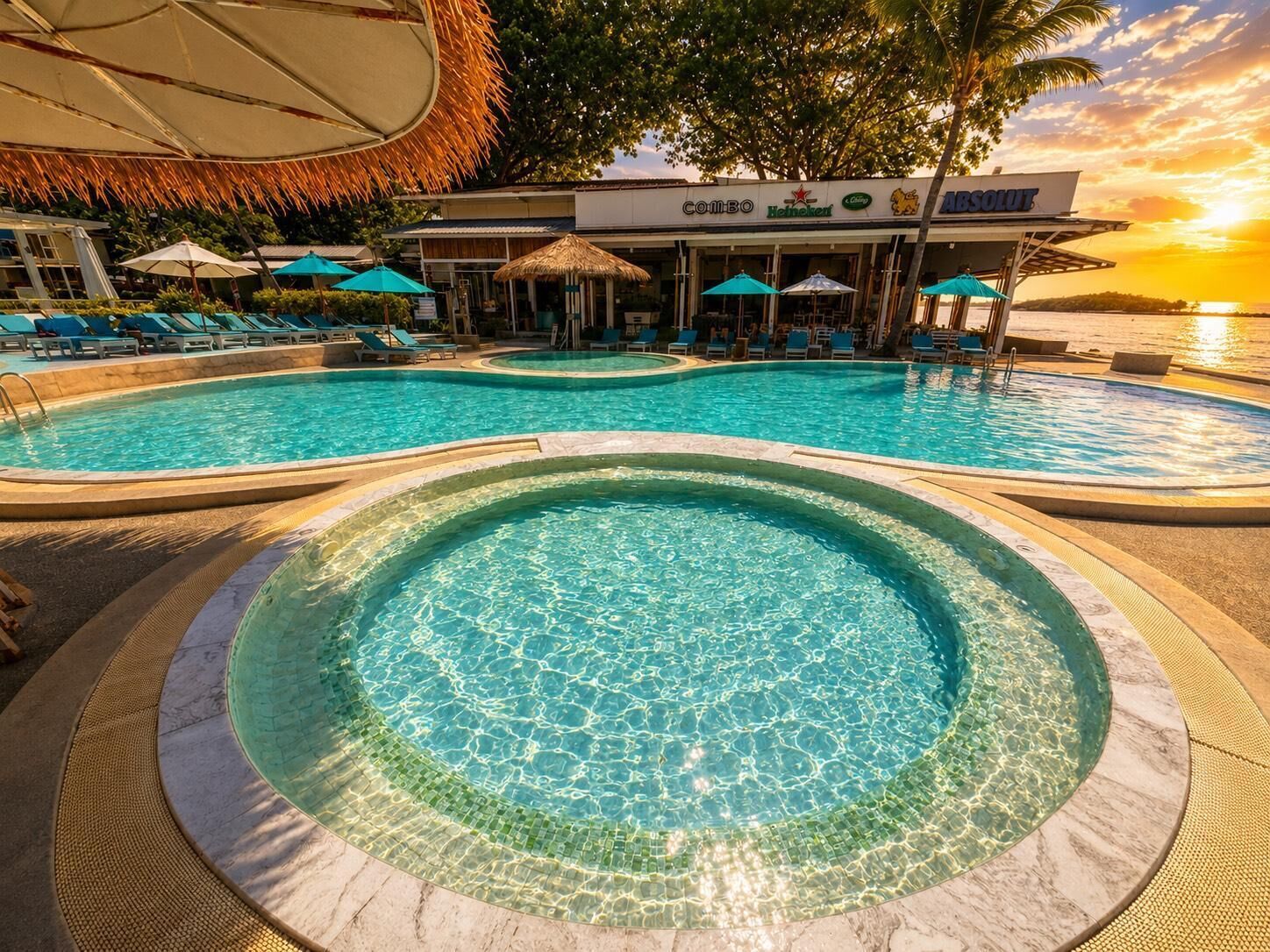 Piscina com cadeiras de sol e guarda-sóis, um bar com anúncios de Heineken e Absolut, e vista para o pôr do sol.