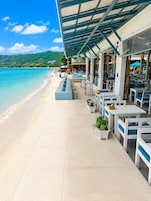 Um restaurante na praia com mesas ao ar livre, piscina e vista para o mar e colinas.