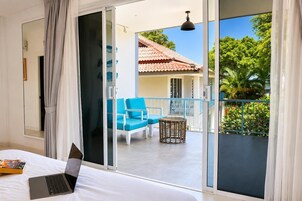 Quarto de hotel com uma cama, um laptop, uma varanda com cadeiras azuis e vista para um edifício com telhado vermelho.
