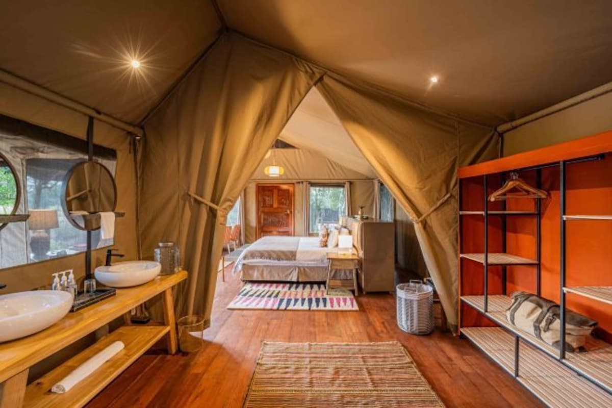 Quarto tipo tenda com um banco de madeira, duas pias, uma cama e uma estante com objetos pendurados.