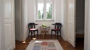 Um corredor com piso de madeira, tapete estampado, duas cadeiras pretas com almofadas vermelhas, uma pequena mesa redonda e um objeto verde, além de uma janela com persianas.