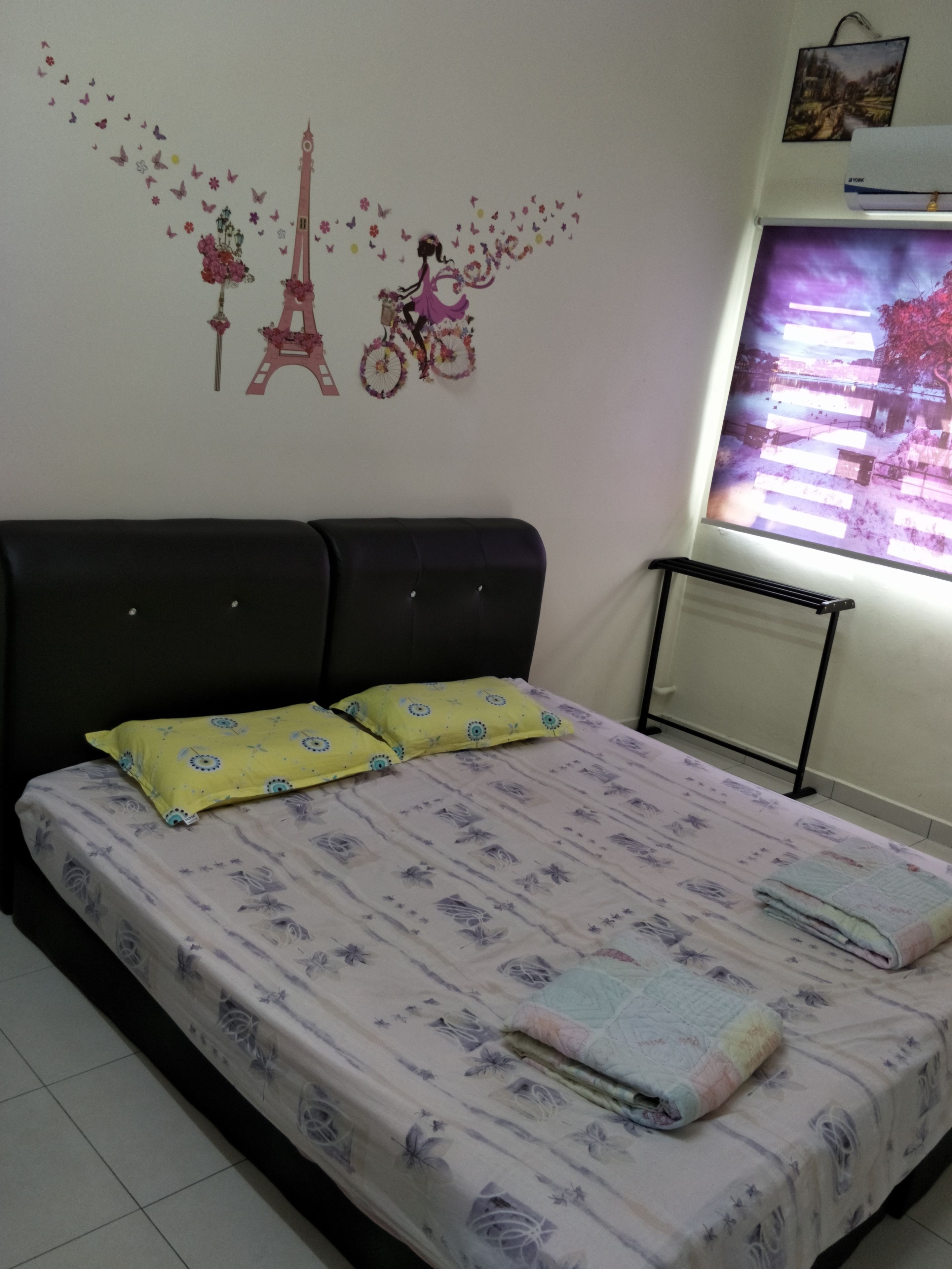 Quarto com cama, duas almofadas, decoração na parede com a Torre Eiffel e uma bicicleta, além de janela com vista para uma ponte.
