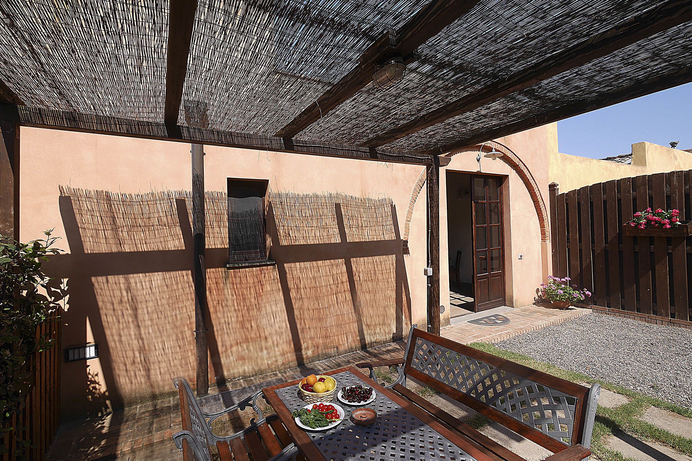Um pátio externo com uma mesa posta para refeição, um banco de madeira e uma pérgola.