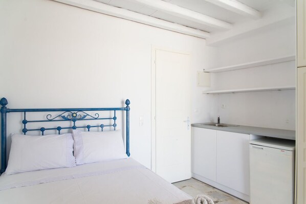 Quarto com estrutura de cama azul de metal, roupa de cama branca, uma porta branca e uma pequena cozinha compacta com pia e armários.