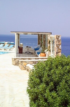 Área de estar ao ar livre com construção em pedra, piscina e espreguiçadeiras, com vista para o mar.