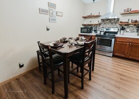 Uma sala de jantar com uma mesa de madeira posta para quatro pessoas, rodeada por cadeiras de madeira escura. Uma cozinha com eletrodomésticos modernos é visível ao fundo.