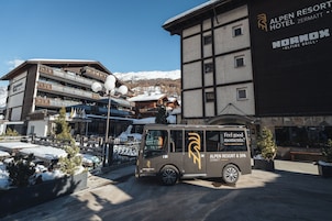 Um ônibus estacionado em frente ao edifício do Alpen Resort & Spa, com vista para montanhas cobertas de neve ao fundo.