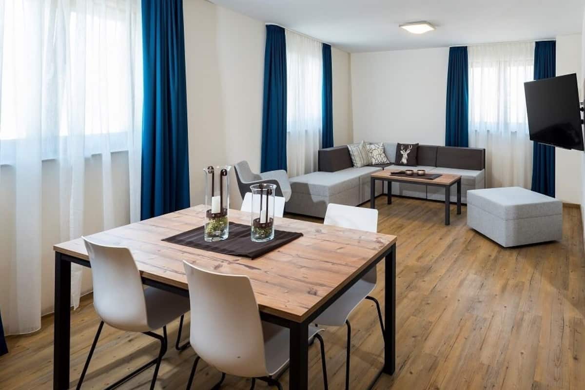 Quarto de hotel moderno com mesa de jantar de madeira, cadeiras brancas, sofá cinzento e televisão de ecrã plano.
