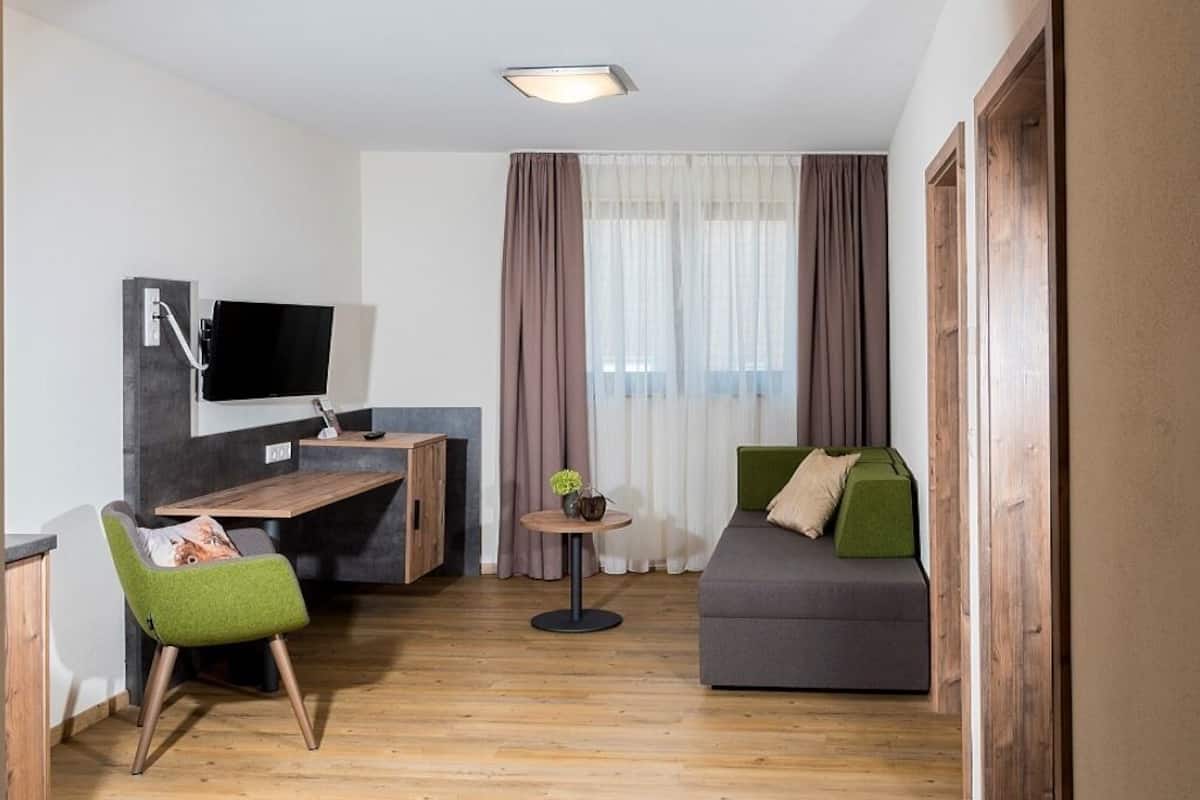 Uma sala de estar moderna com piso de madeira, uma poltrona verde, um sofá cinza, uma pequena mesa redonda e uma TV fixada na parede.