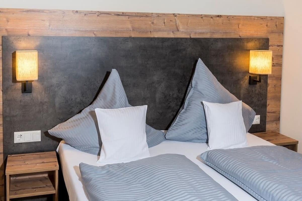 Uma cama de casal com cabeceira cinzenta e duas luminárias laterais.
