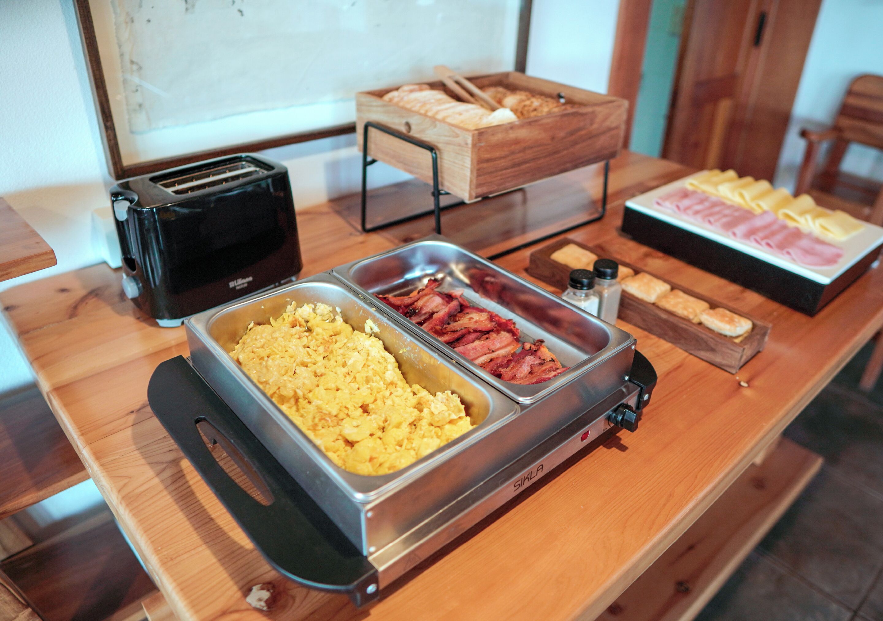 Buffet com ovos mexidos, bacon e diversos pães.