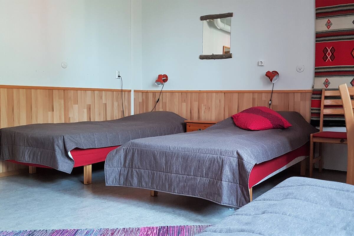 Quarto com duas camas de solteiro, cabeceira de madeira, um espelho pequeno, travesseiro vermelho, uma cadeira e um tapete estampado.
