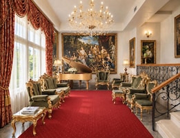 Uma sala imponente com carpete vermelho, um piano de cauda, cadeiras ornamentadas e um grande quadro clássico.