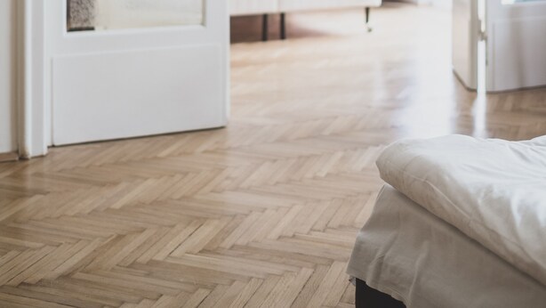 Um quarto com piso de madeira em padrão herringbone, uma cama com roupa de cama branca e uma janela grande com vista para o exterior.