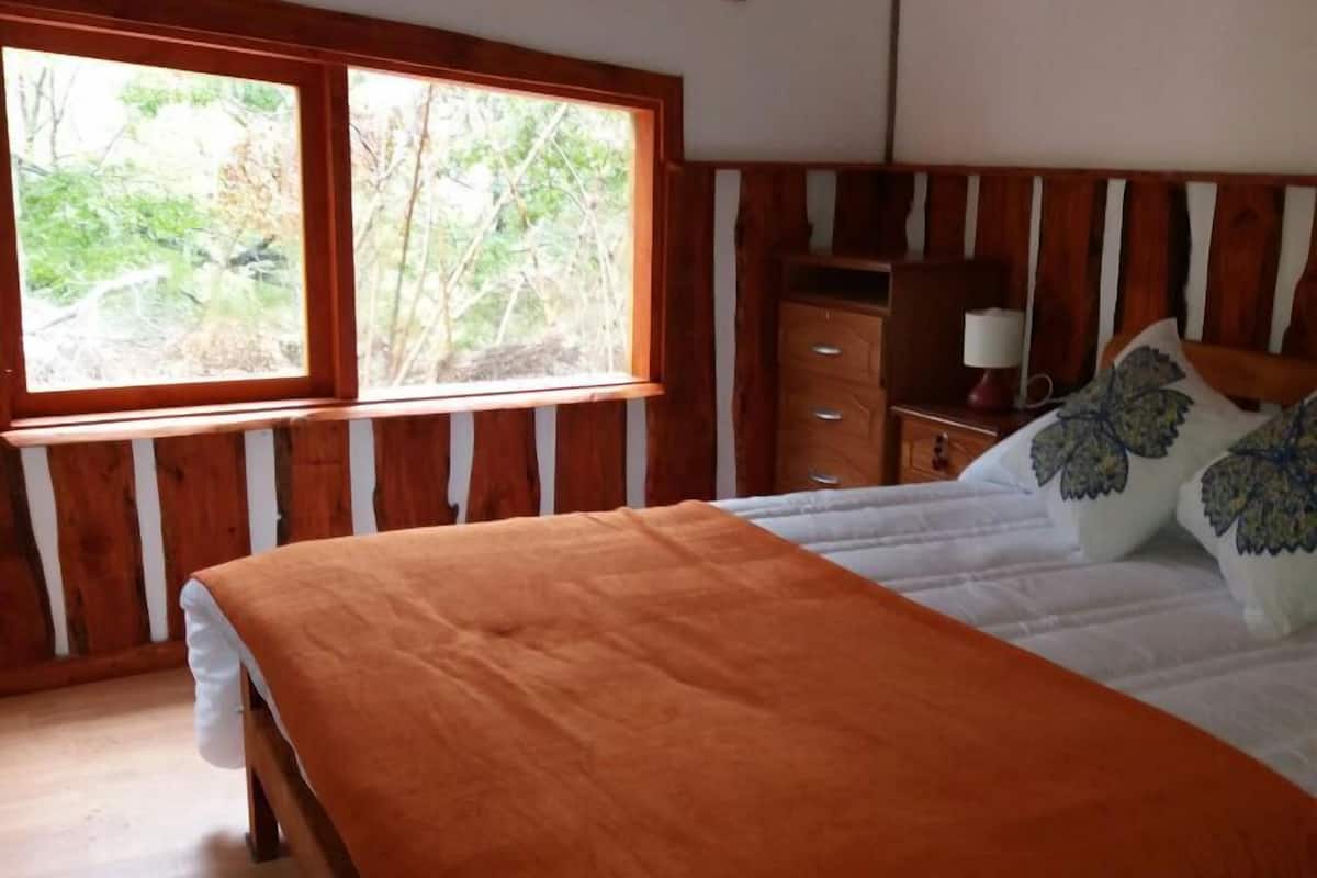 Quarto com cabeceira de madeira, uma janela grande, cama com colcha laranja e mesa de cabeceira com abajur.