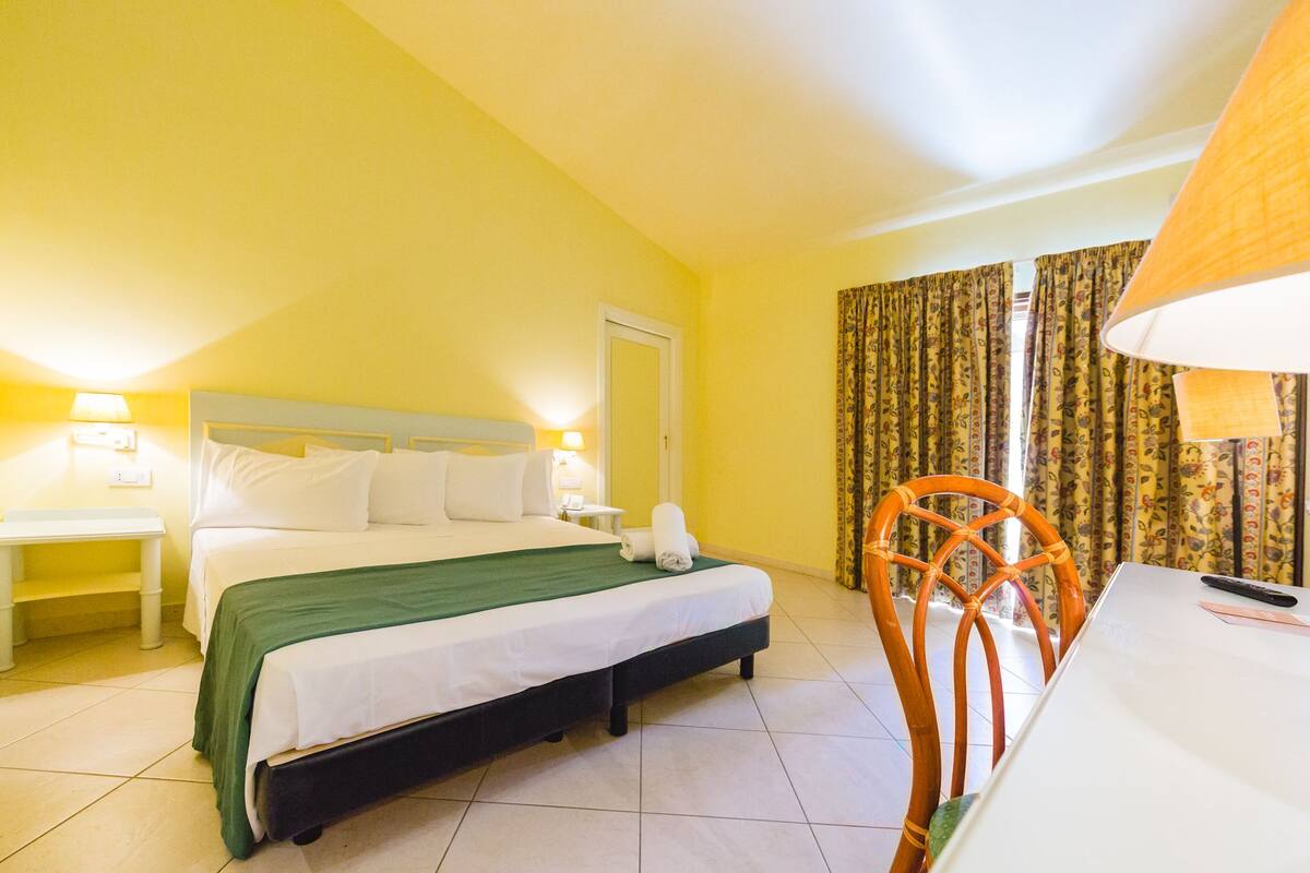 Quarto de hotel com uma cama, uma cadeira, uma escrivaninha e cortinas.