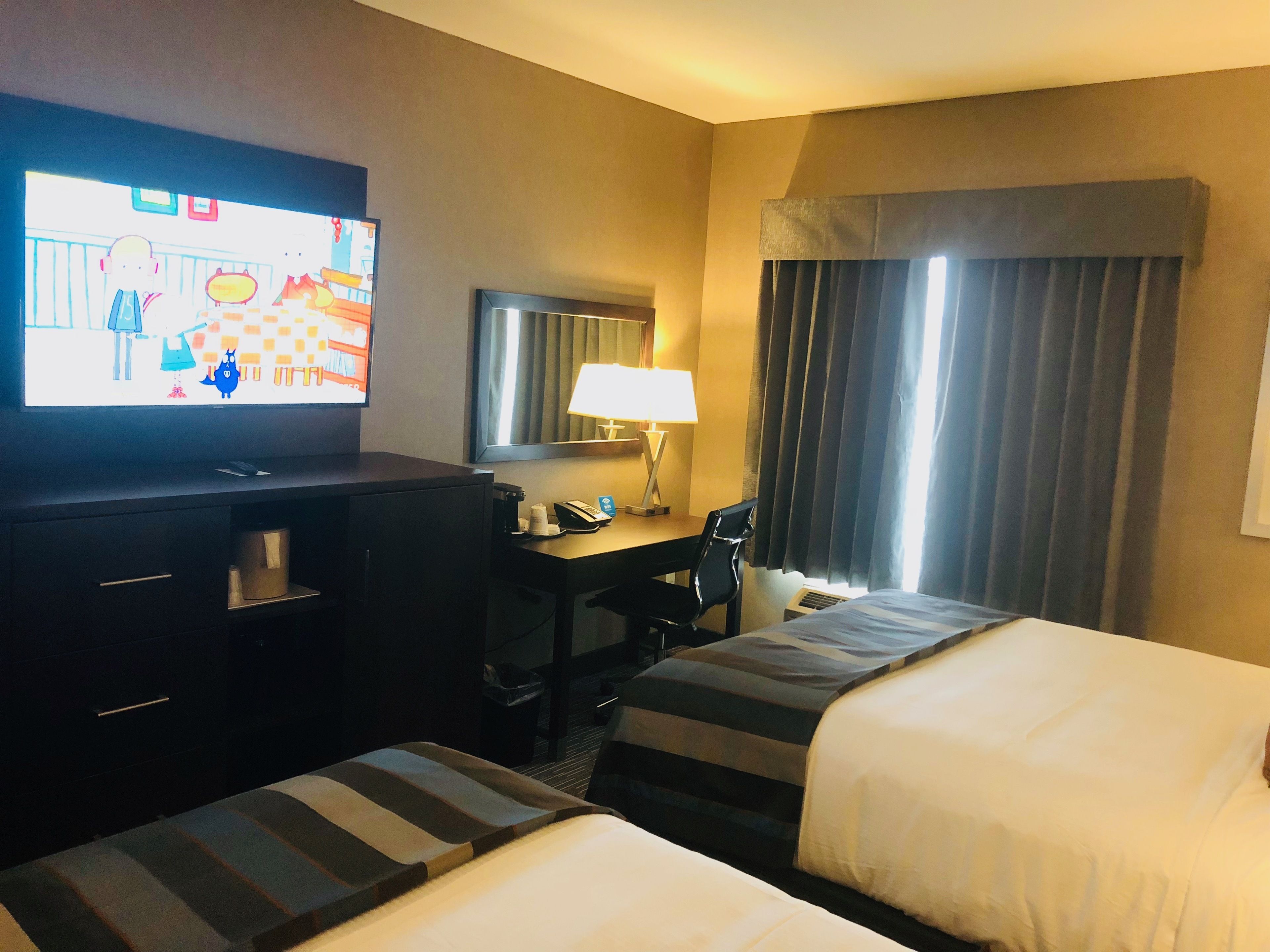 Quarto de hotel com televisão, escrivaninha com luminária, duas camas e janela com cortinas.