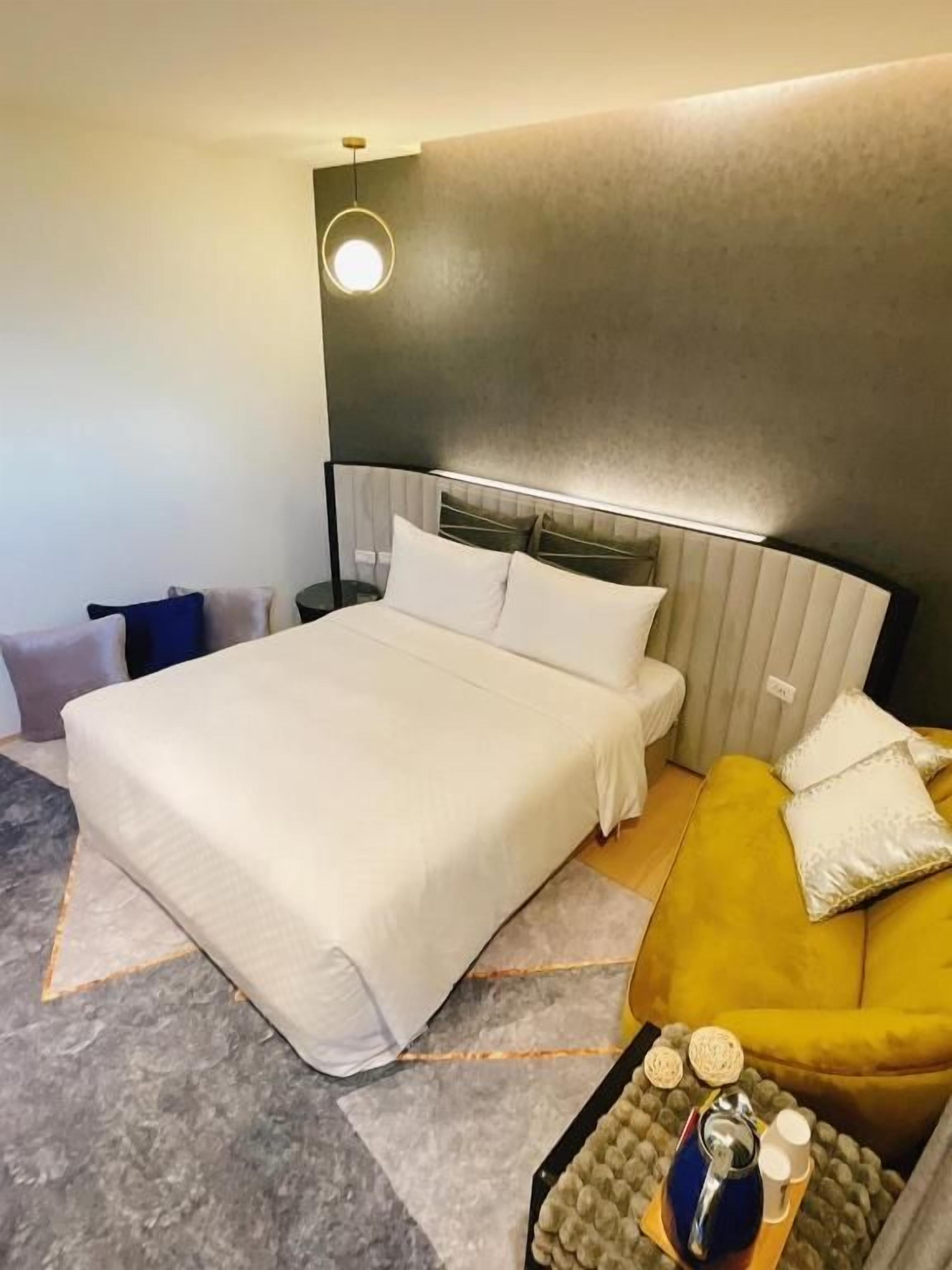 Quarto de hotel com cama, mesa de cabeceira, abajur, sofá e bandeja com objetos.