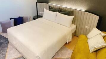 Quarto de hotel com cama, mesa de cabeceira, abajur, sofá e bandeja com objetos.