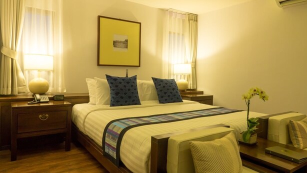 Quarto de hotel com uma cama grande, duas mesas de cabeceira com abajures, telefone ao lado da cama e um quadro na parede, além de uma pequena mesa com uma planta.