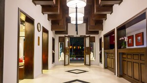 Um saguão de hotel com uma porta aberta no centro que dá para a rua, um candelabro e um chão estampado.