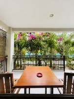 Uma mesa de madeira com quatro cadeiras, um objeto vermelho sobre a mesa e vista para um jardim com flores cor-de-rosa e um corrimão preto.