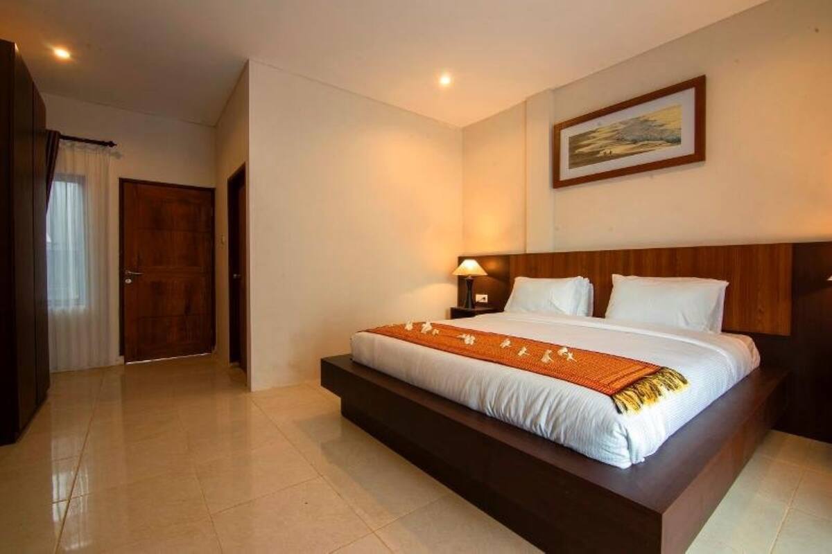 Quarto de hotel com uma cama grande, cabeceira de madeira, mesa de cabeceira com abajur, quadro pendurado na parede e porta com cortina.
