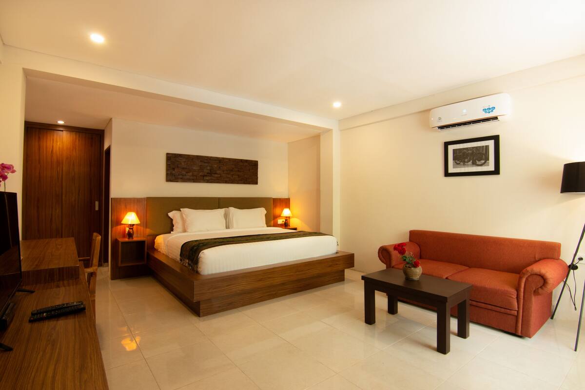 Quarto de hotel com uma cama, um sofá vermelho, uma mesa de centro, uma televisão e um ar condicionado.