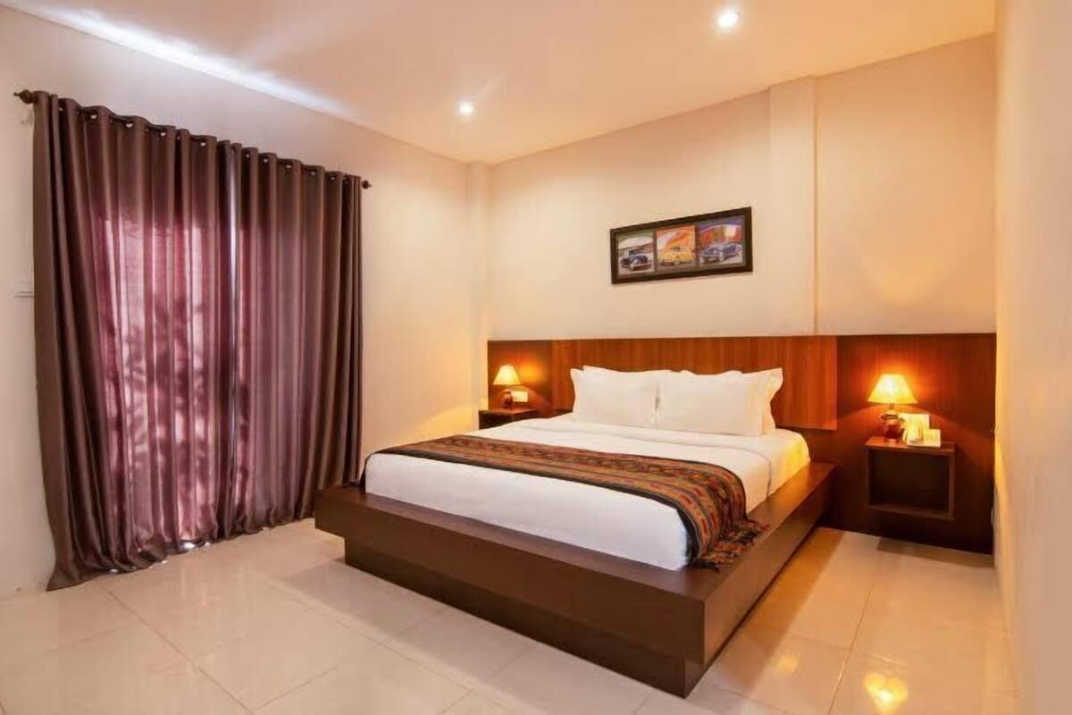 Quarto de hotel com uma cama grande, duas mesas de cabeceira com abajures, cabeceira de madeira e um quadro na parede.
