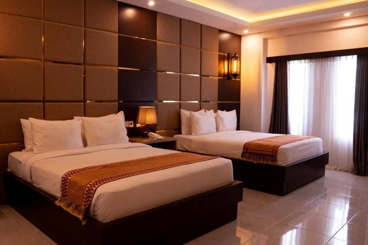 Quarto de hotel com duas camas, uma cabeceira grande e acolchoada, abajures e uma janela ampla com cortinas.