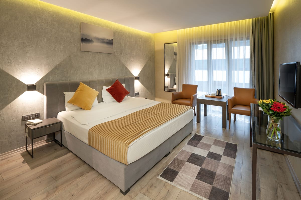 Quarto de hotel com uma cama grande, abajures, televisão de tela plana, uma mesinha com um vaso de flores e uma cadeira.