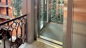 Um elevador com porta de vidro, guarda-corpo de metal e vista para um edifício com varandas e vegetação.