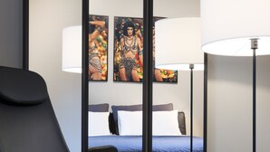 Quarto de hotel moderno com uma poltrona azul, uma cama com colcha azul, um espelho grande e um abajur.