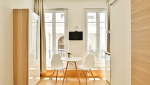 Quarto moderno com uma mesa redonda e duas cadeiras brancas, uma televisão de tela plana na parede e grandes janelas com vista para edifícios.