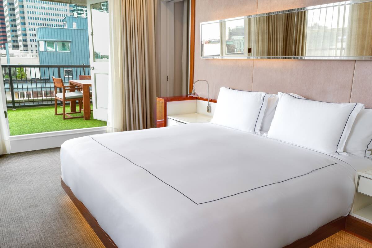 Quarto de hotel com uma cama grande, uma escrivaninha e uma cadeira. Tem uma varanda com vista para edifícios.