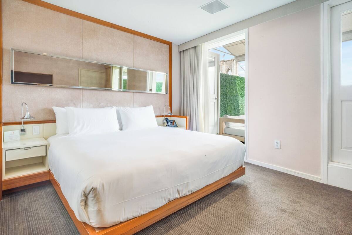 Uma cama grande com estrutura de madeira, uma mesa de cabeceira com abajur, um espelho sobre a cama e vista para um conjunto de arbustos verdes através de uma porta de vidro.