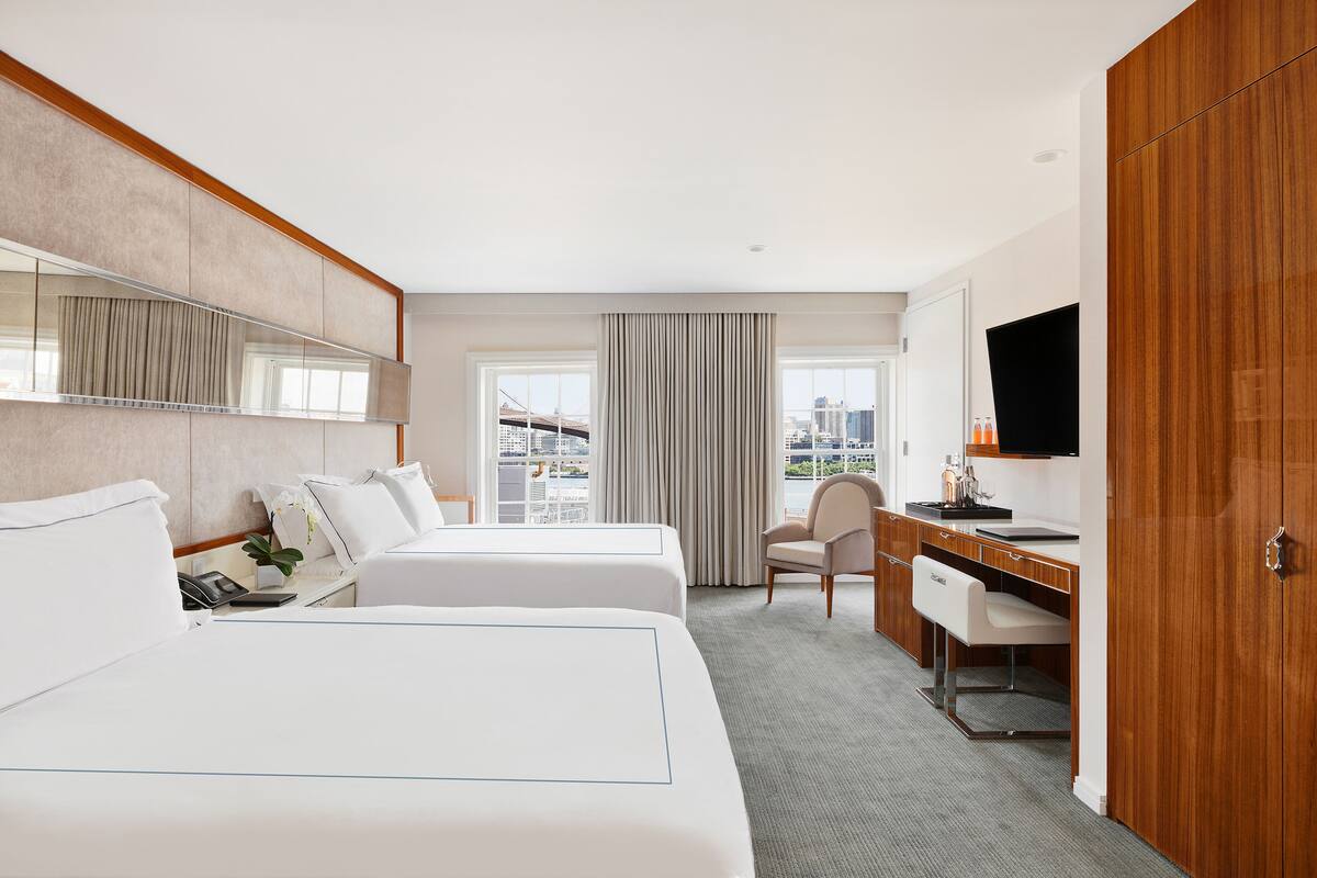 Quarto de hotel com duas camas, uma escrivaninha, uma cadeira, televisão e vista para a cidade.
