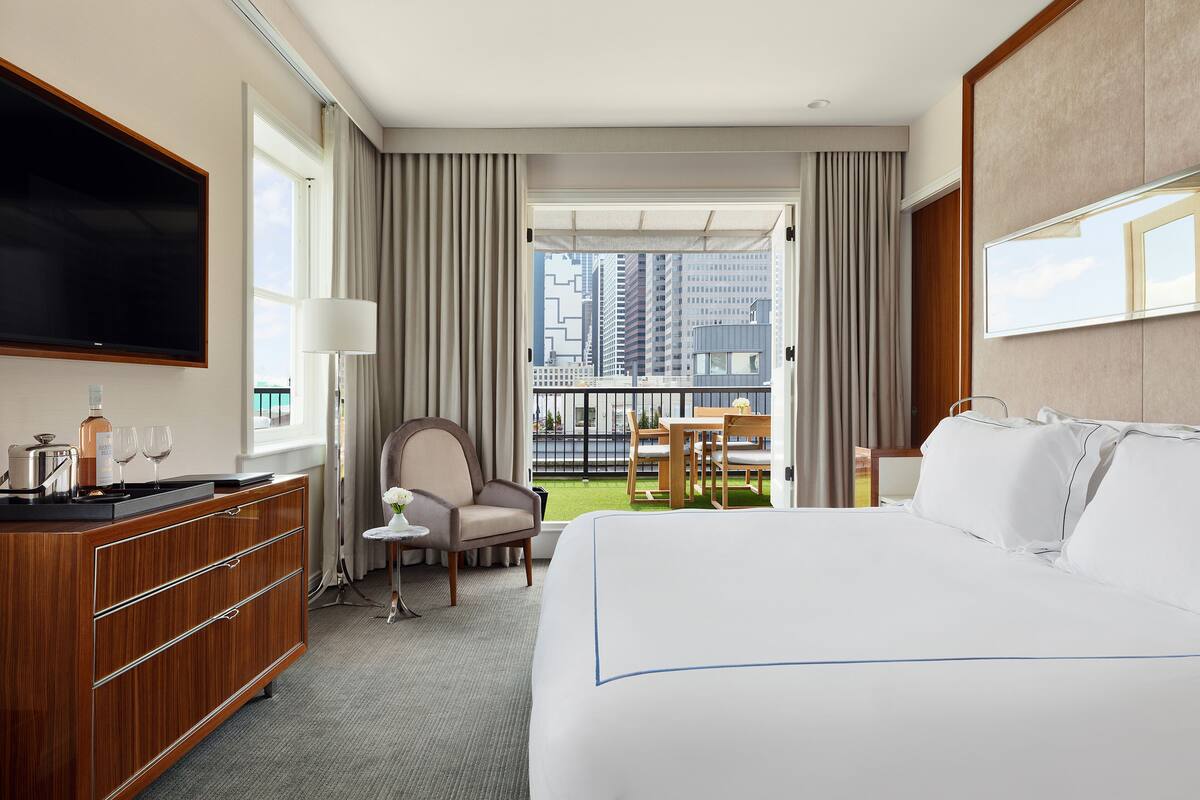 Quarto de hotel com uma cama grande, penteadeira de madeira, televisão de tela plana, área de jantar com mesa e cadeiras, e vista para a paisagem urbana pela janela.
