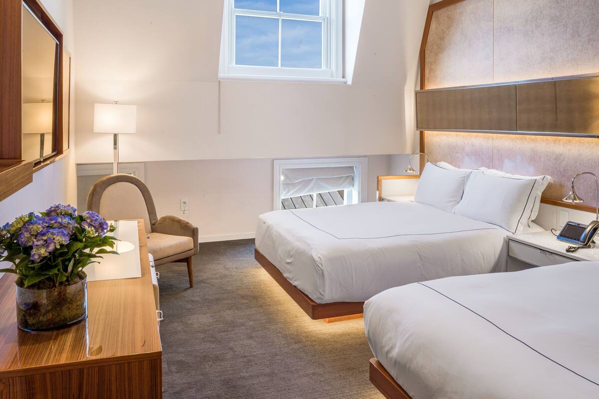 Quarto de hotel com duas camas, uma escrivaninha, uma cadeira, mesa de cabeceira, luminária e janela com vista para o céu.