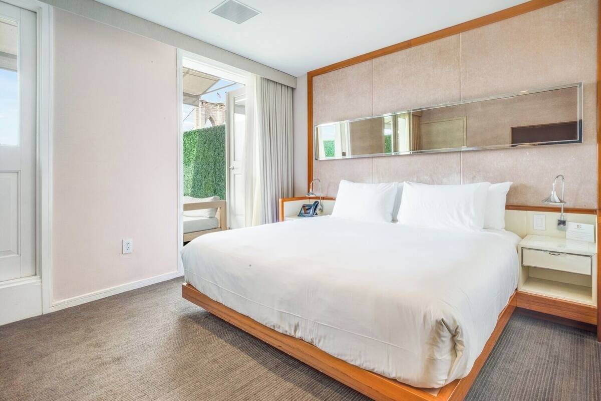 Quarto de hotel com uma cama grande, uma mesa de cabeceira, um espelho e vista para o exterior.