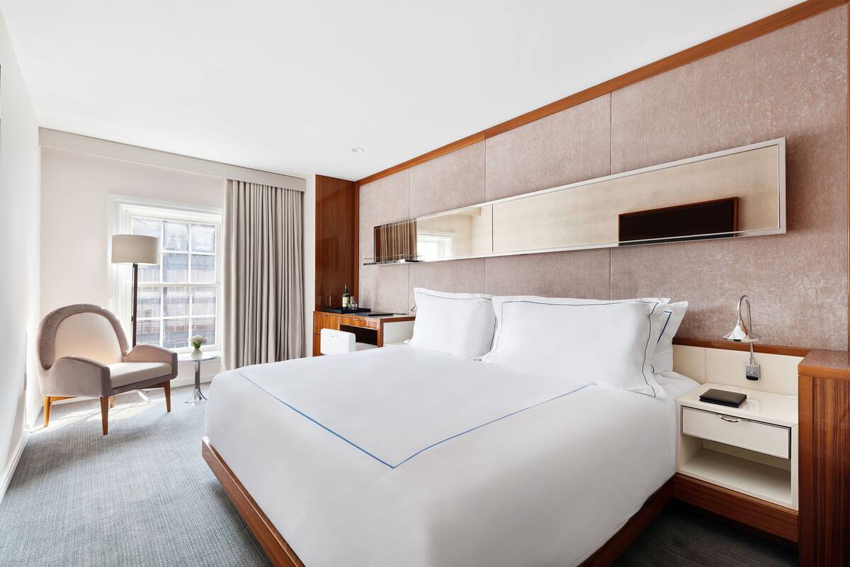 Quarto de hotel com uma cama grande, uma escrivaninha com luminária, uma cadeira e uma janela com cortinas.
